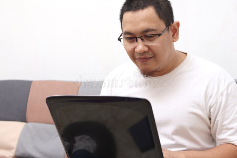 Cheerful Asian Man Using Computer and Smiling while Sitting on the Sofa ...