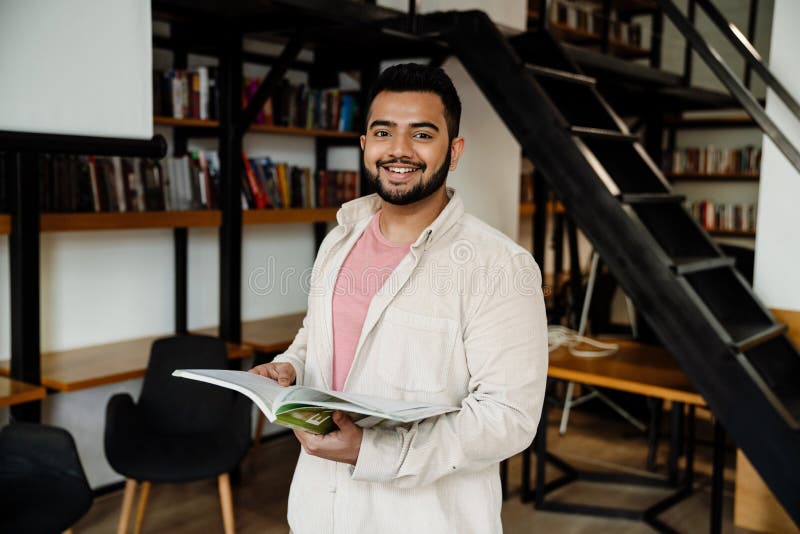 Cheerful Indian Man Holding Textbook while Standing in Library Stock ...
