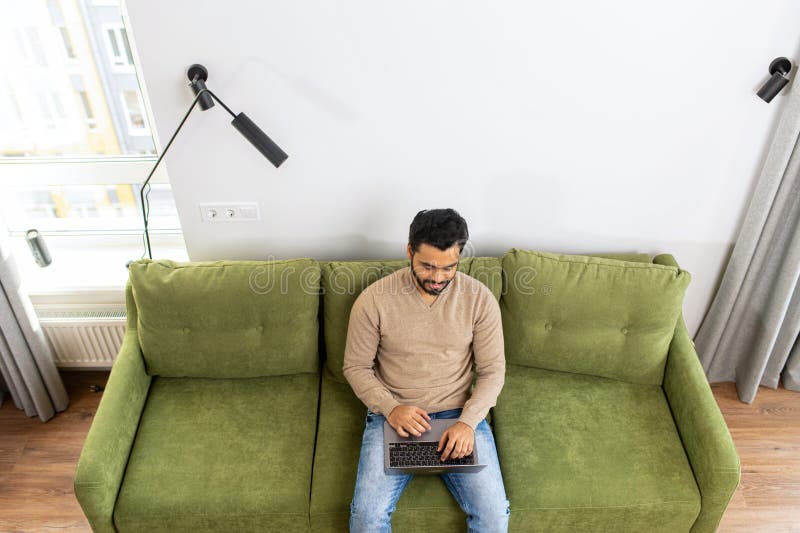 Cheerful Indian Man Wearing Casual Wear Sitting on the Sofa and Using ... Cheerful Indian Man Wearing Casual Wear Sitting on the Sofa and Using ...