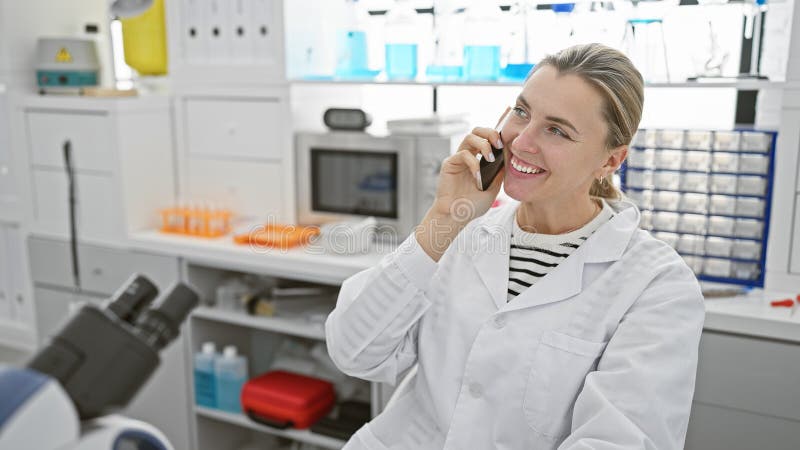 A Cheerful Woman Scientist in a Lab Coat Talking on a Phone in a Bright ...