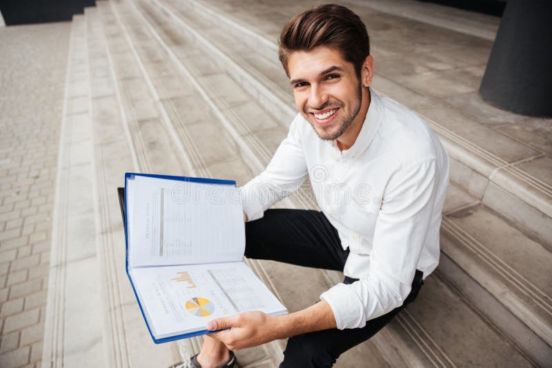 Cheerful Young Businessman Sitting and Reading Business Plan in Folder ...