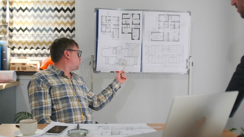 Client and Contractor Finalize Plans at Desk with Floorplans on Board, Seated Professional ...