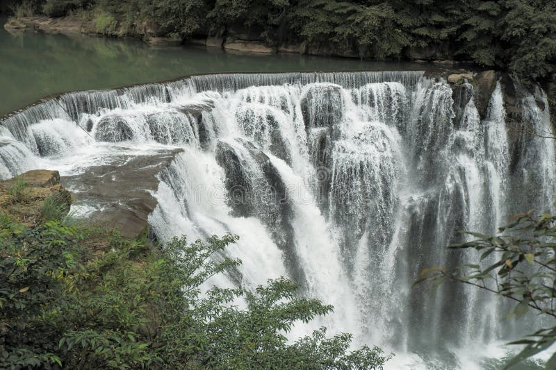 Waterfall Flowing Over Rocky Cliff with Green Trees and River in ...