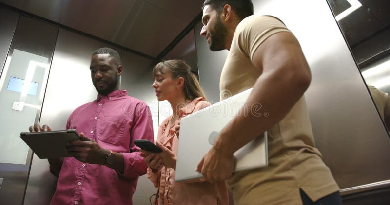 Collaborating in Elevator, Diverse Colleagues Using Tablet, Smartphone, and Laptop for Work ...