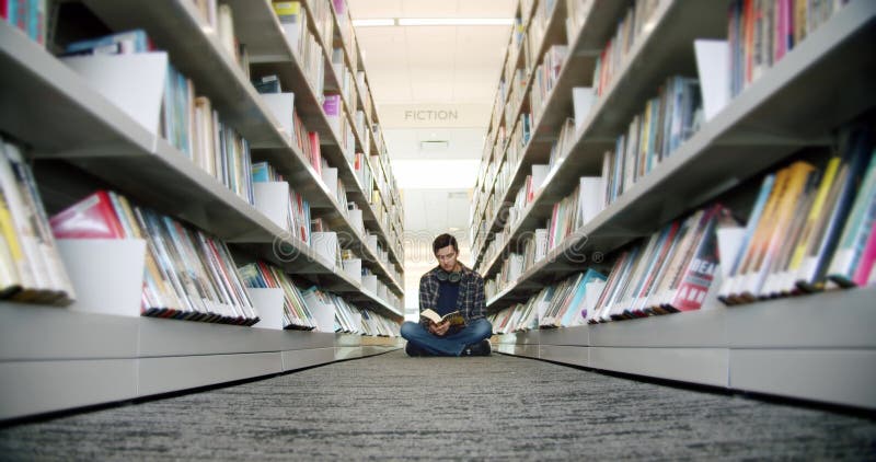 College Student Sitting on the Floor in the Library and Reading a Book ...