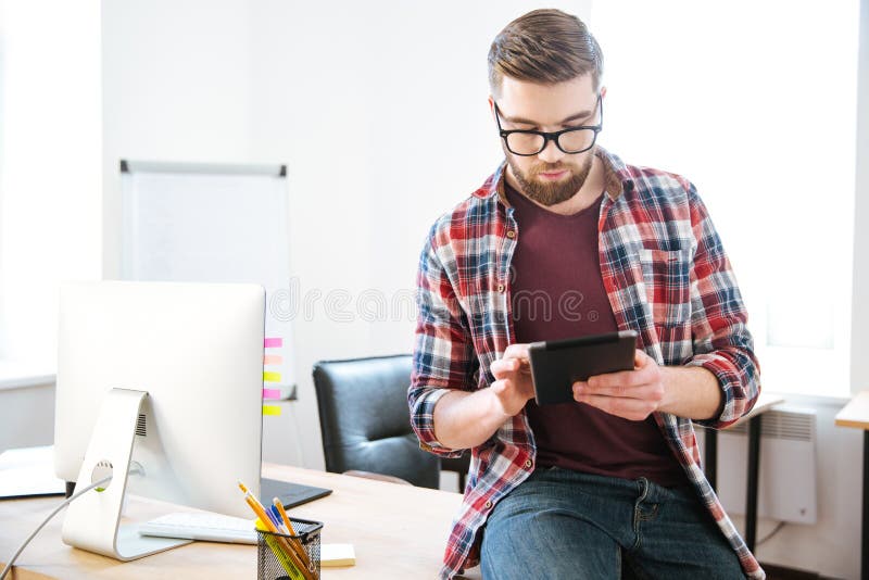 Concentrated man sitting on table in office and using tablet - Stock ...