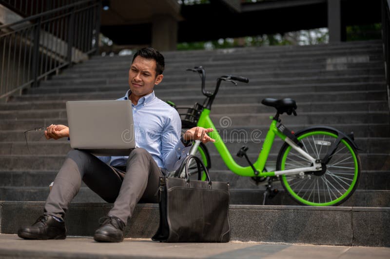 A Confused Asian Businessman Sits on Steps with a Laptop, Showing ...