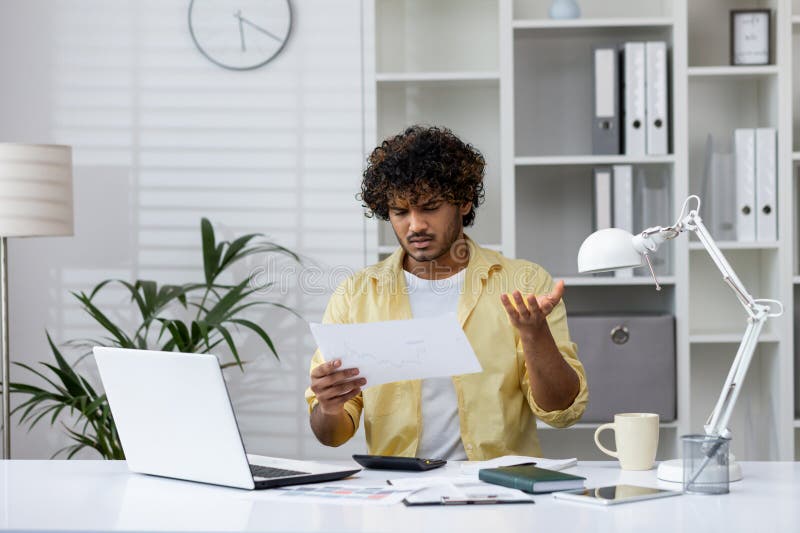 Confused Man Examining Document in Modern Office Setting with Laptop ...