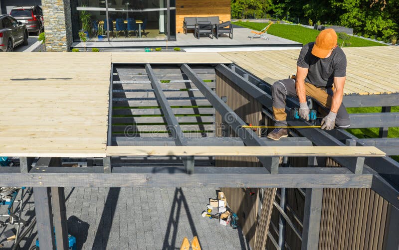 Construction of a Wooden Deck on a Modern House in Daylight with a Worker Measuring the Planks ...