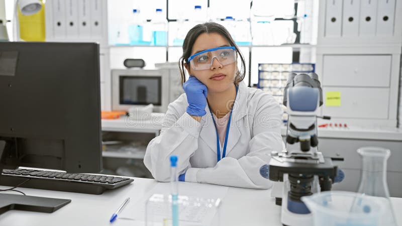 A Contemplative Hispanic Woman Scientist in Lab Coat and Safety Glasses ...