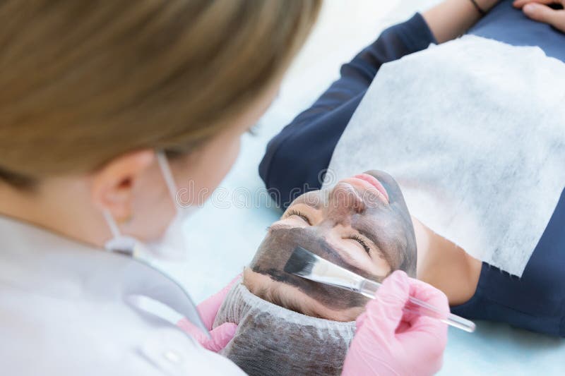The Cosmetologist In Pink Gloves With A Brush Applies A Carbon Mask For