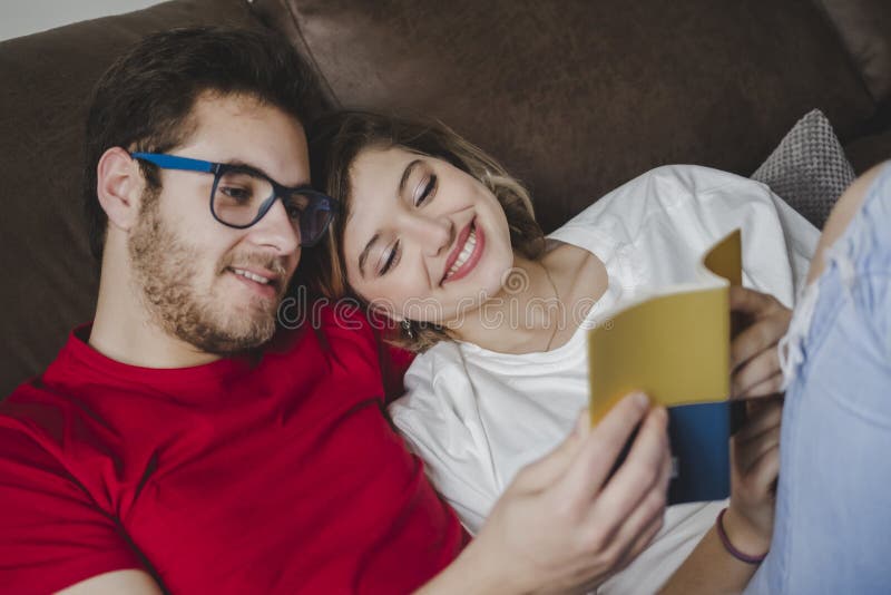 Smiling Couple Reading Book While Resting On Sofa In Living Room At