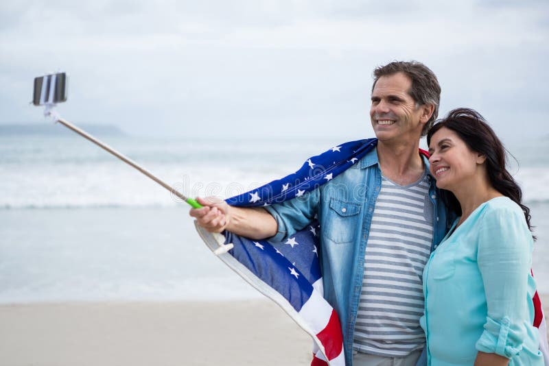 Couple Wrapped In American Flag Taking Selfie On Beach Stock Image