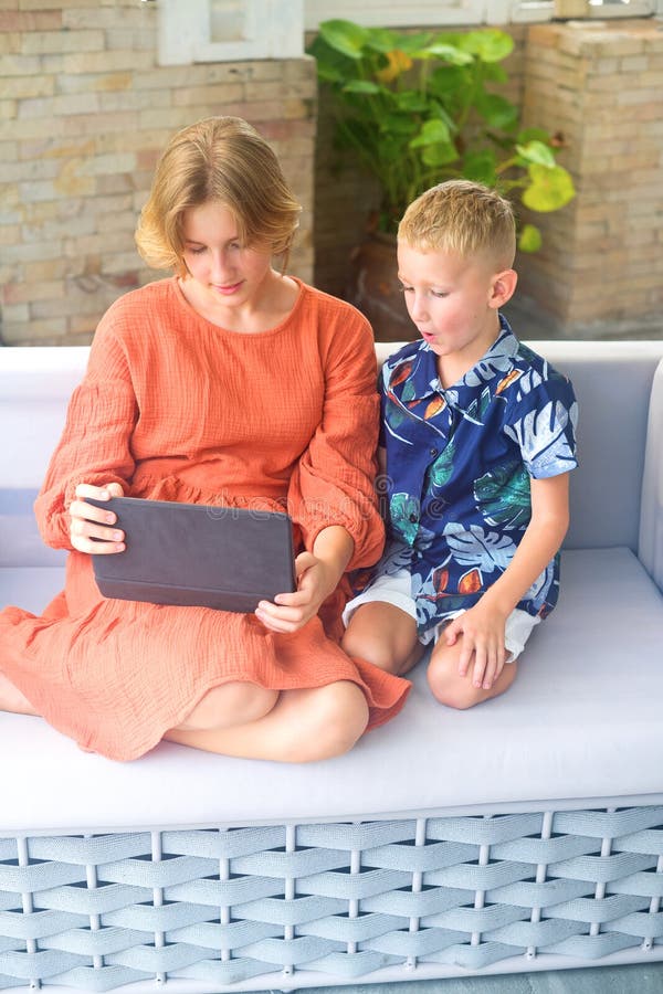 Cute Siblings Brother And Sister Sitting On Sofa At Home And Playing On Wireless Tablet Together