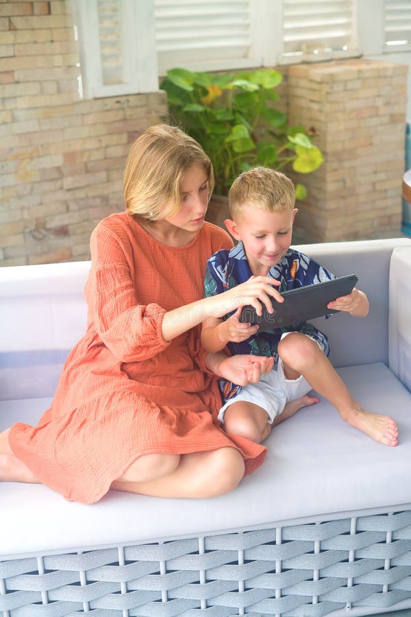 Cute Siblings Brother And Sister Sitting On Sofa At Home And Playing On Wireless Tablet Together