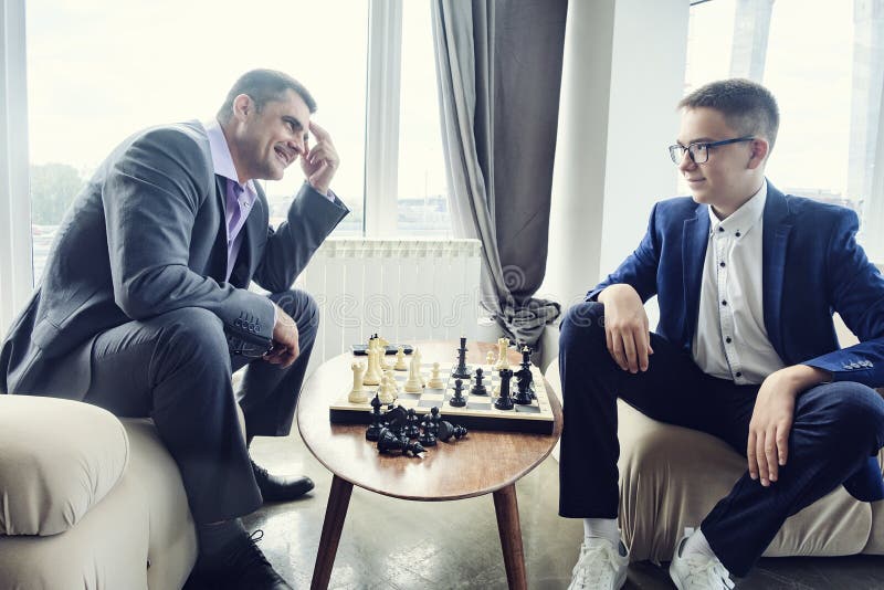 Dad And Son Are Sitting At A Table By A Wide Window And Playing Chess Making Moves With Pieces