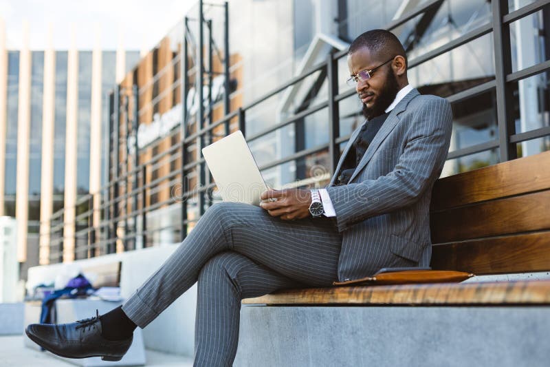 A Dark-skinned Man in a Business Suit is Using a Laptop Outdoors ...