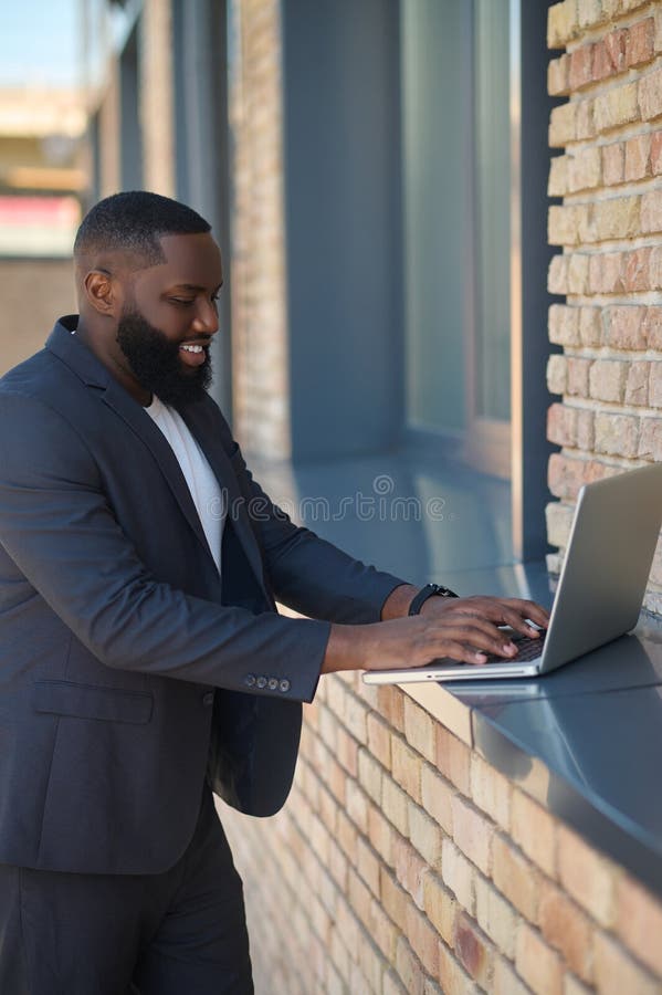 A Dark-skinned Man Staning Outside and Typing Something on a Laptop ...