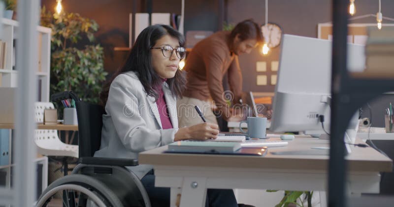 Disabled Young Lady in Wheelchair Working in Office Looking at Computer Screen and Writing in ...