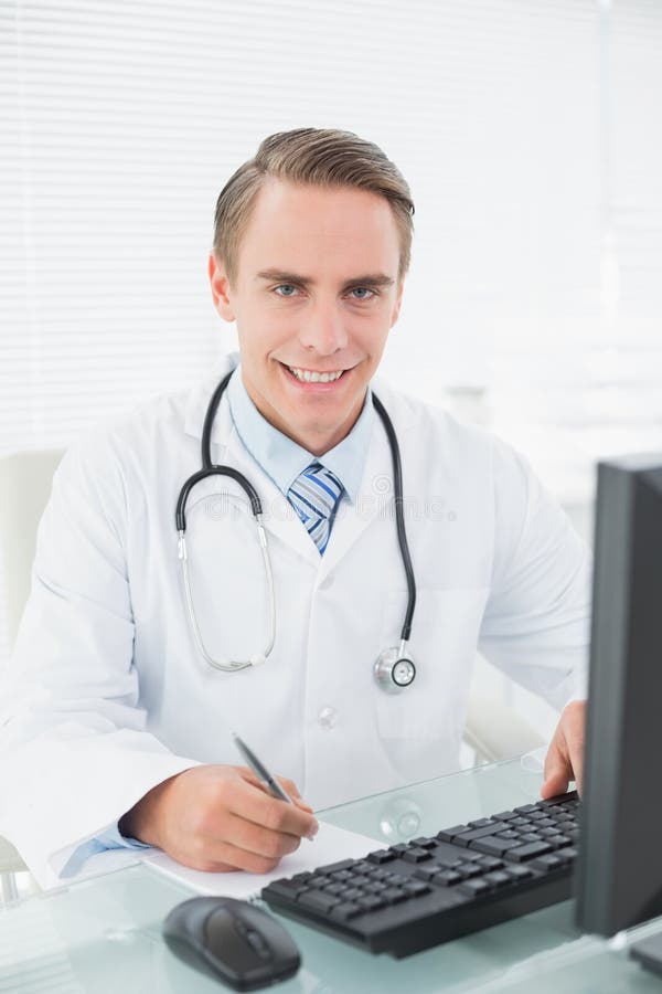 Doctor Writing a Note while Using Computer at Medical Office Stock ...
