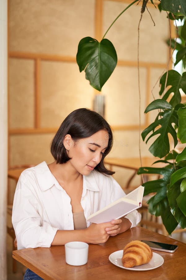 Dreamy Young Smiling Asian Woman Reading Book Sitting In Cafe Eating