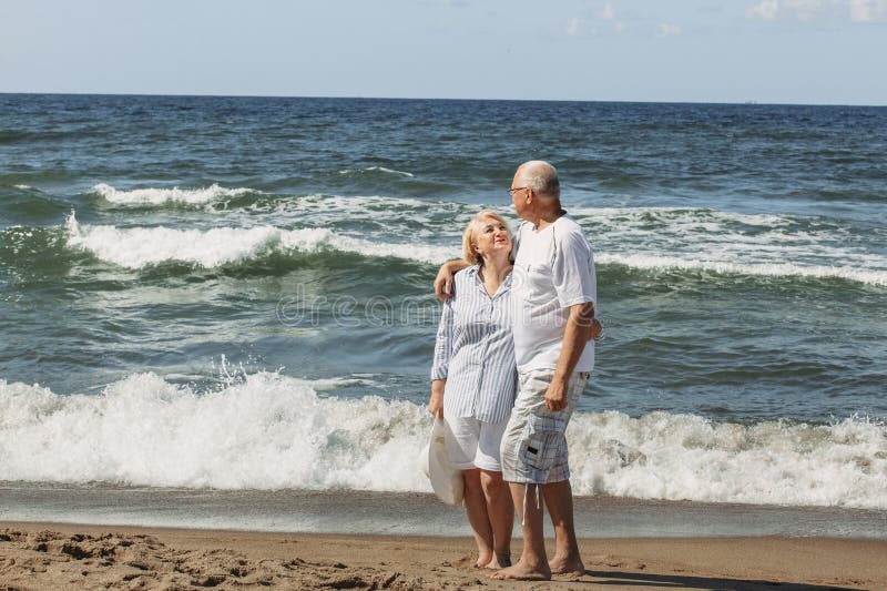 An Elderly Couple Stand Hugging On The Beach By The Sea Look At Each