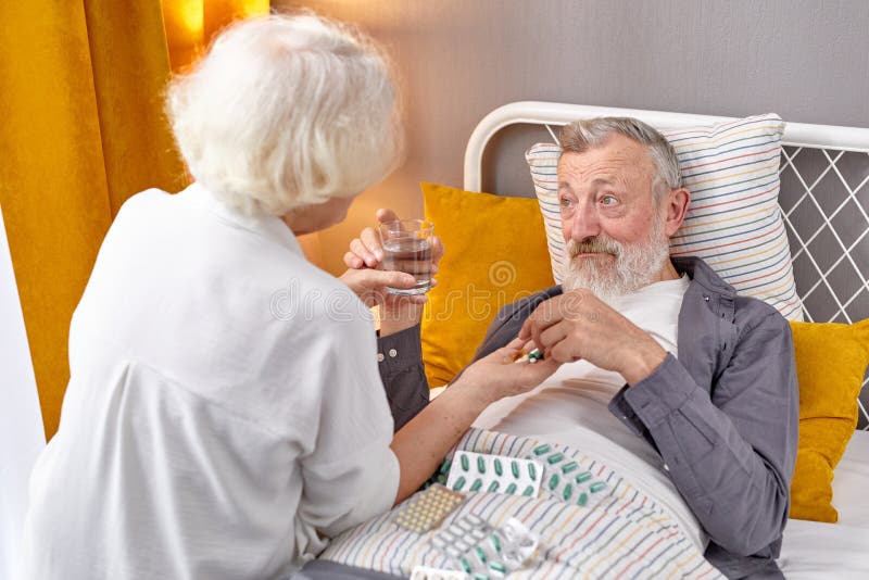 Elderly Man Taking Medicines And Drinking Water While Lie On Bed Wife