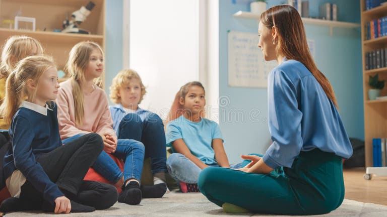 Elementary School Creativity Class: Children Sitting on Carpet while Caring Teacher Explains ...