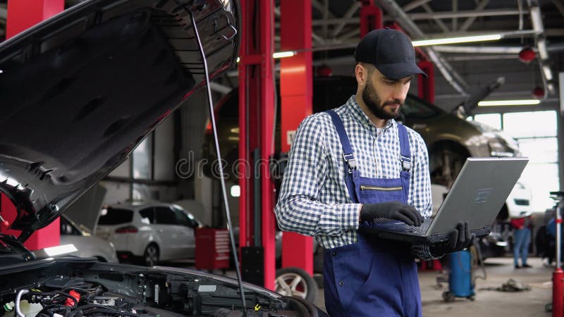 Experienced Mechanic in Uniform is Using a Laptop while Repairing Car ...