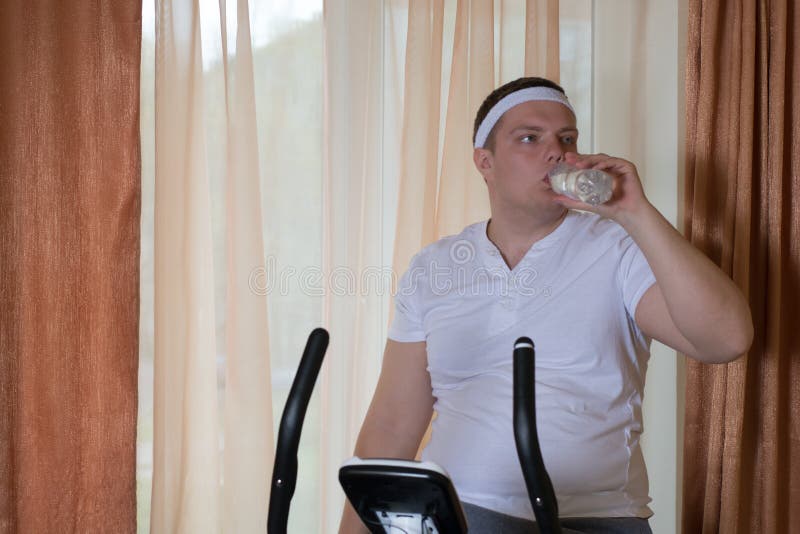 Fat Guy Exercising on Stationary Training Bicycle and Drinking Water ...