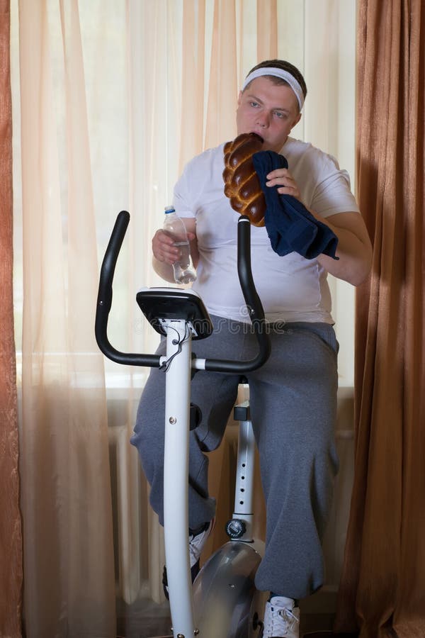 Fat Guy Exercising on Stationary Training Bicycle and Eating a Bun ...
