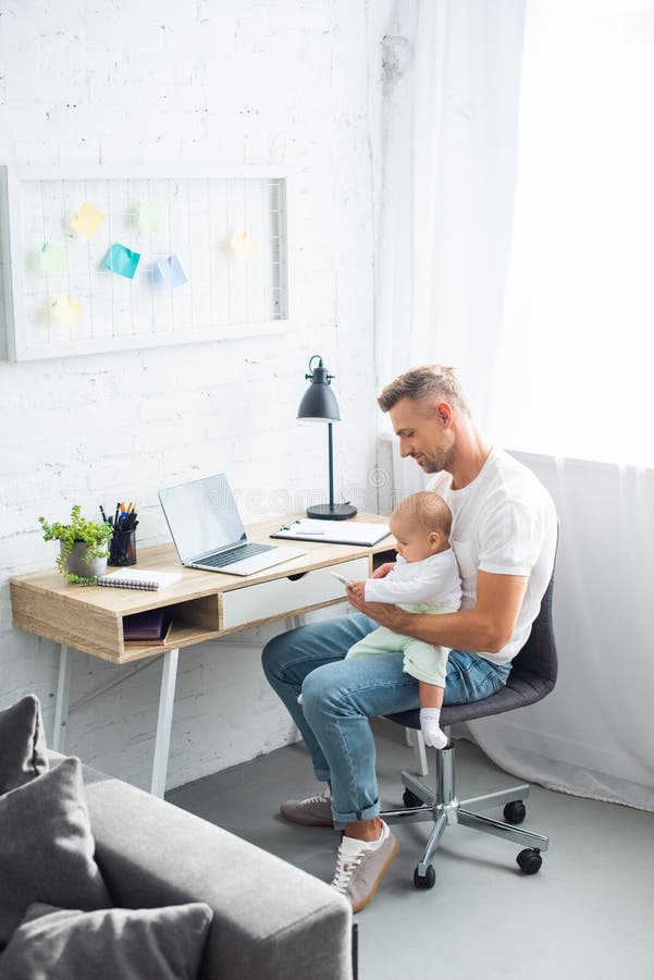 Father Sitting at Computer Desk, Using Smartphone and Holding Baby ...