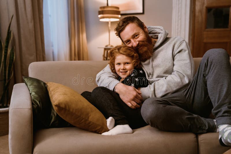 Father And Son Hugging And Smiling At Camera While Resting On Sofa