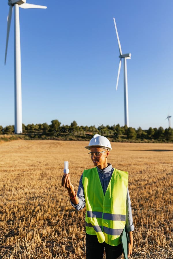 Female Engineer Holding an Energy-saving Light Bulb Against Windmills ...