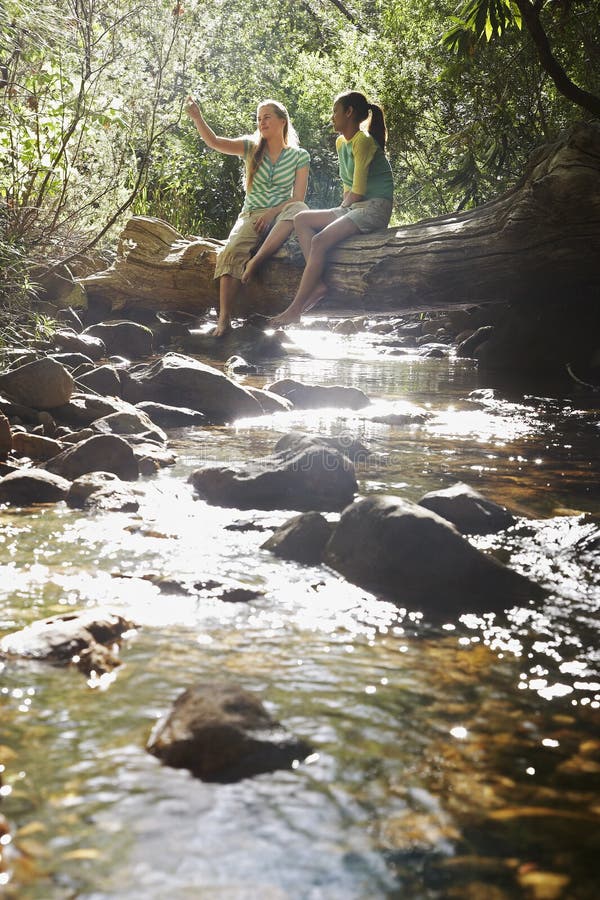 Female Friends Sitting on Log Over Forest Stream Stock Image - Image of ...