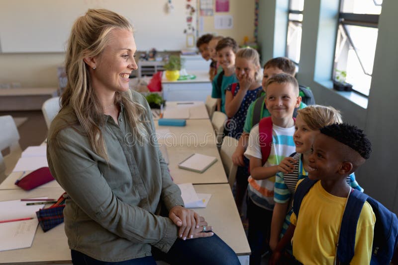 Female School Teacher Sitting On A Desk In A Classroom With A Group Of
