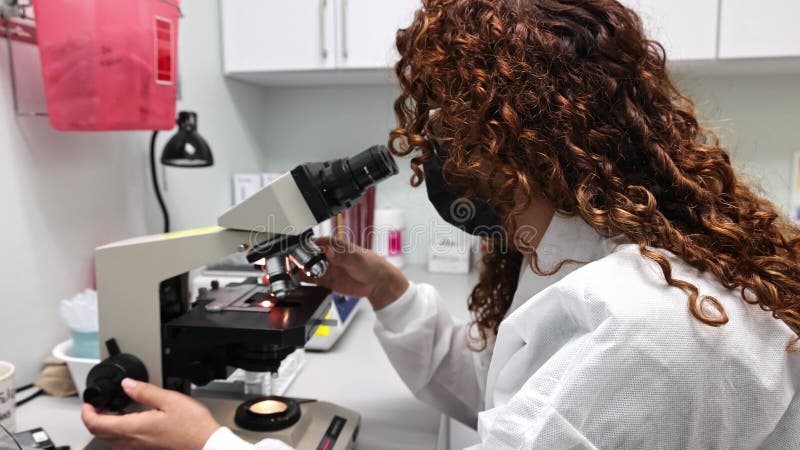Female Scientist in a Lab Coat and Mask Examines a Slide Sample through ...