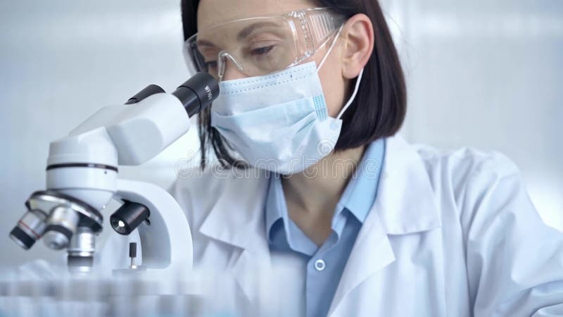 Female Scientist in Lab Goggles and Protective Mask Using Microscope in ...