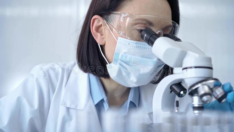 Female Scientist in Lab Goggles and Protective Mask Using Microscope in ...
