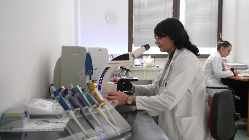 Female Scientist Wearing a Lab Coat Examines Samples Under a Microscope ...