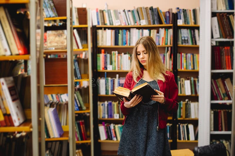 Female Student Reading a Book between Bookshelves in University Library ...