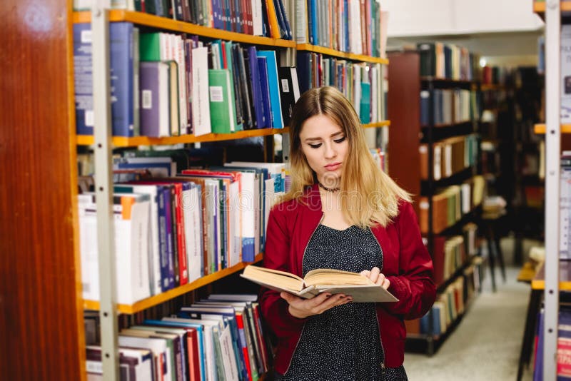 Female Student Reading a Book between Bookshelves in University Library ...