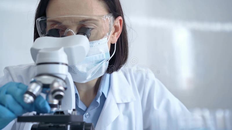 Female Scientist in Lab Goggles and Protective Mask Using Microscope in ...