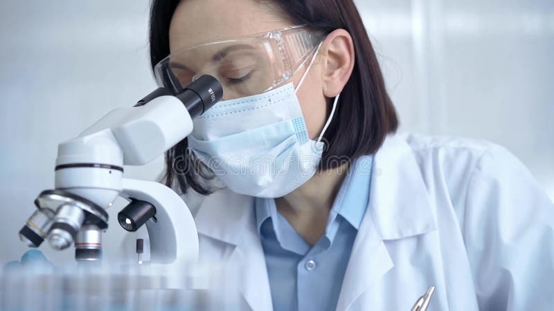 Female Scientist in Lab Goggles and Protective Mask Using Microscope in ...