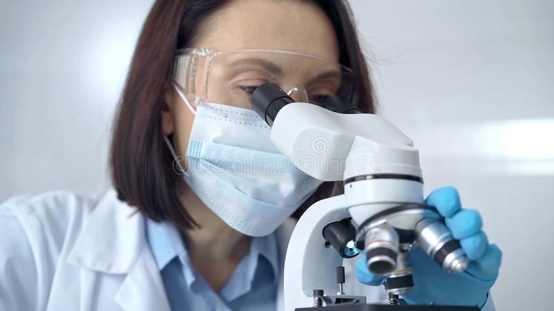 Female Scientist in Lab Goggles and Protective Mask Using Microscope in ...