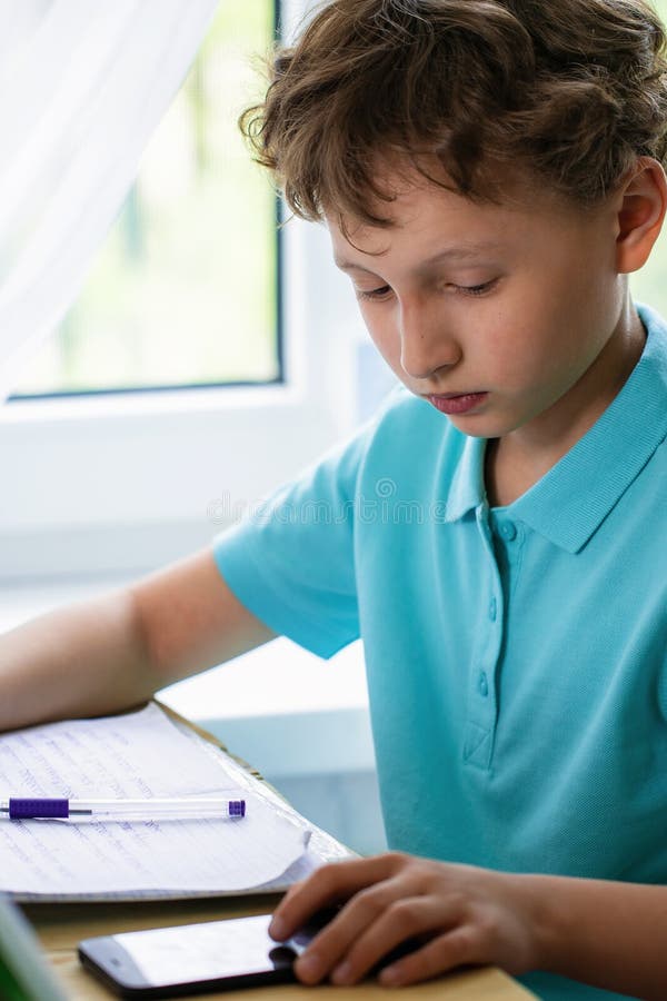 Focused Boy Sits at a Table and Does Homework Using a Smartphone Stock ...