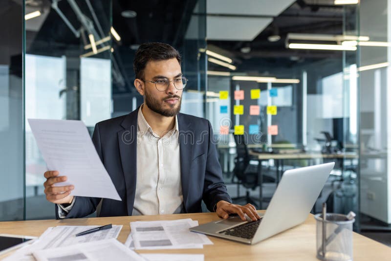 A Focused Businessman at His Desk Reviews Paperwork while Using His ...