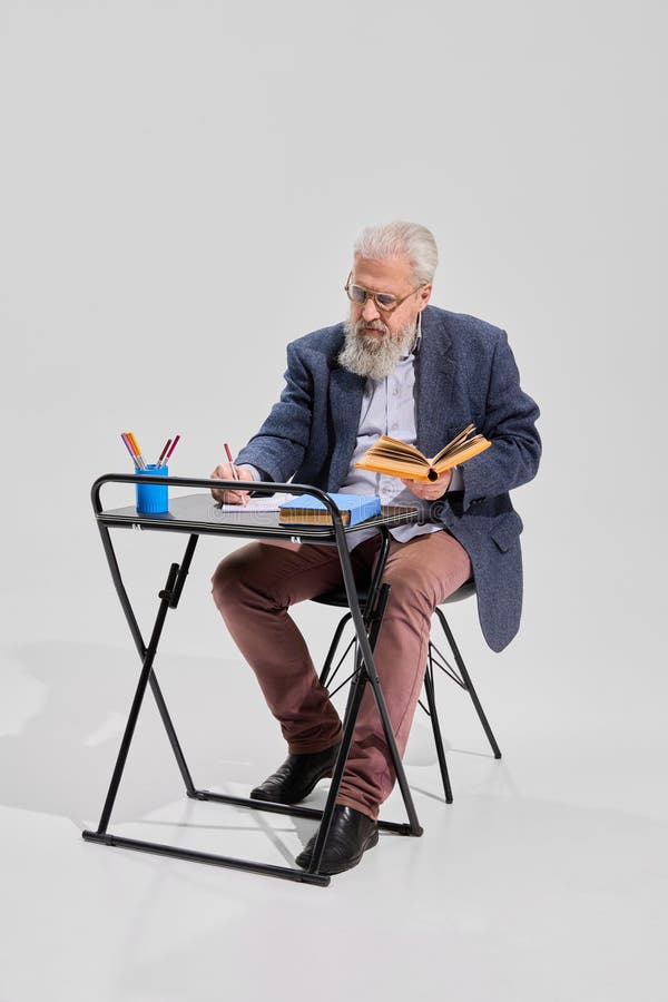 Focused Elderly Man Sitting at Desk, Holding Open Book and Making Notes ...