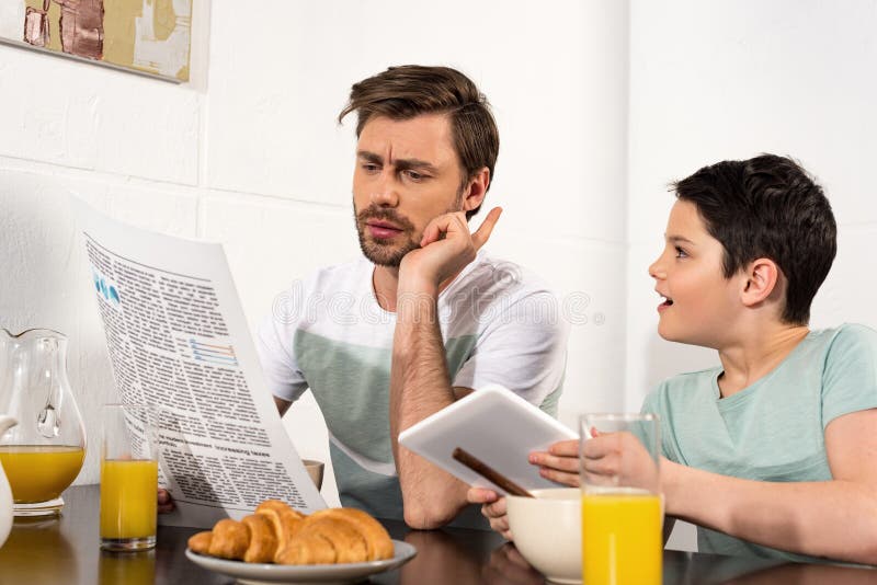 Father Reading Newspaper and Son Using Digital Tablet during Breakfast ...