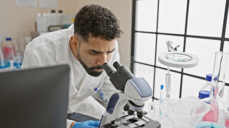 A Focused Hispanic Man with a Beard Examines Samples Using a Microscope ...